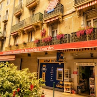 Um edifício com varandas decoradas com flores, um letreiro com 'La Résidence Berceau De Bernadette Lourdes' e um caixa eletrônico no andar térreo.