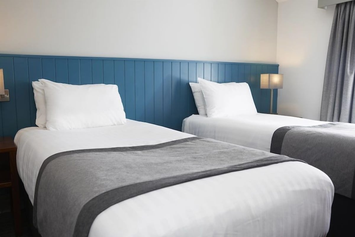 Quarto de hotel com duas camas, cada uma com roupa de cama branca e um cobertor cinza.