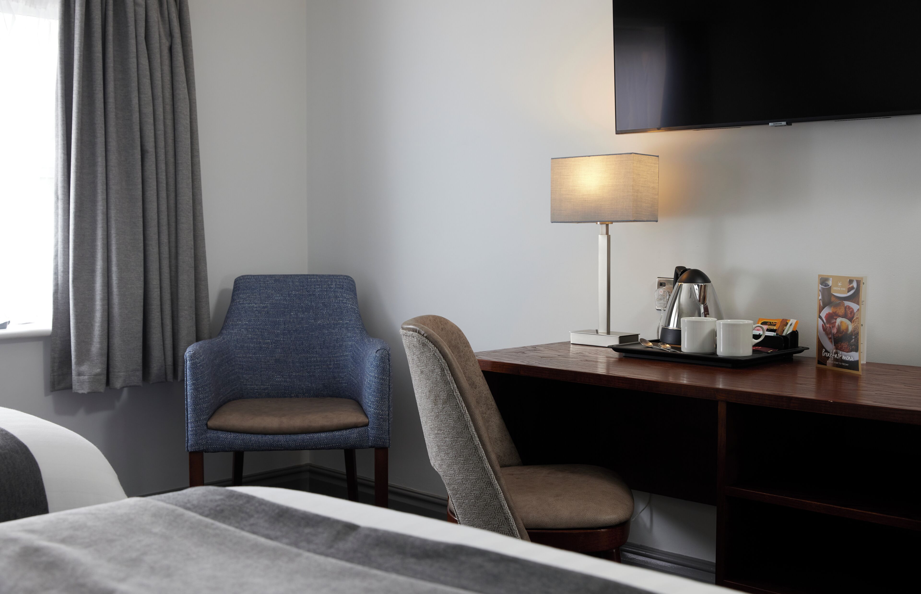 Quarto de hotel com uma cama, uma escrivaninha com abajur, uma cadeira, uma televisão e uma janela com cortinas.