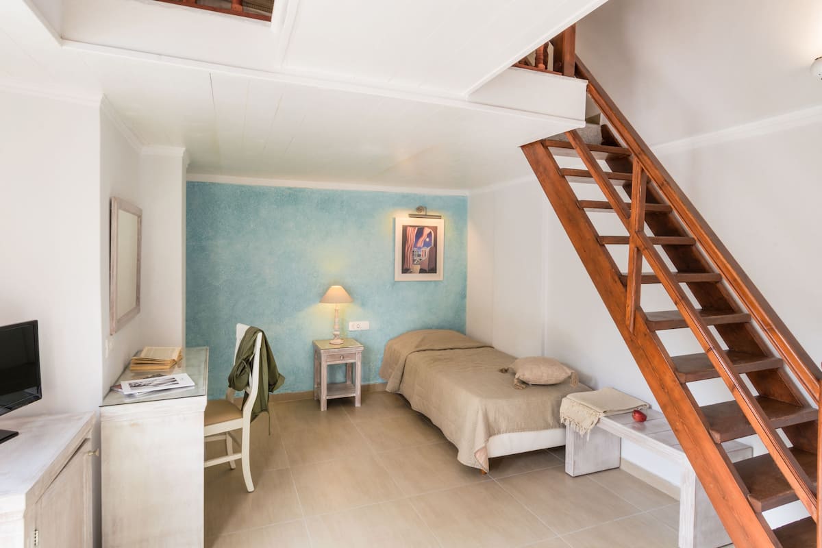 Quarto com cama, escrivaninha, cadeira, mesa pequena com abajur e escada de madeira.