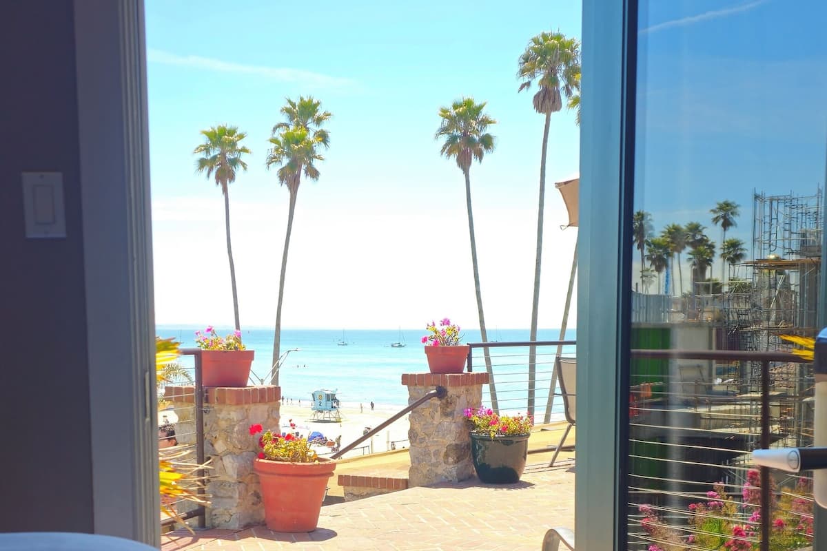 Quarto com vista para a praia, palmeiras e área externa com plantas em vasos.