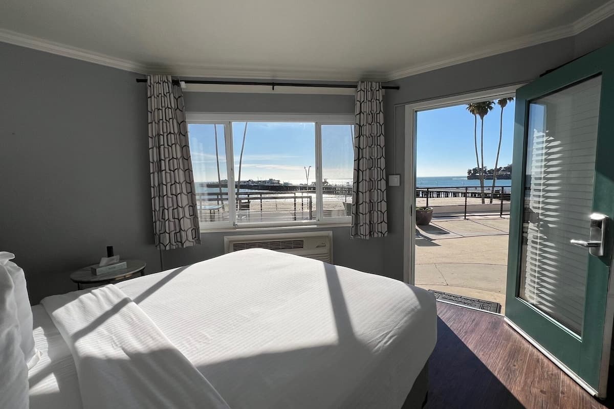 Quarto de hotel com cama, janela com cortinas e vista para a praia, além de porta para o exterior.