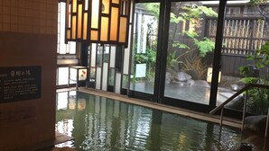 Uma onsen japonesa tradicional, com banheira de pedra, teto de madeira e vista para um jardim através de grandes portas de vidro.