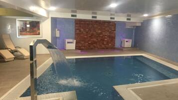 Piscina interna com fonte de água, cadeiras de descanso e parede revestida com padrão de tijolos.
