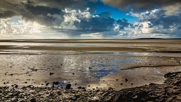 Uma praia com uma extensa faixa de areia molhada e um primeiro plano rochoso.