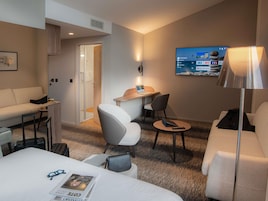 Quarto de hotel com cama, escrivaninha, poltrona, sofá e televisão na parede.