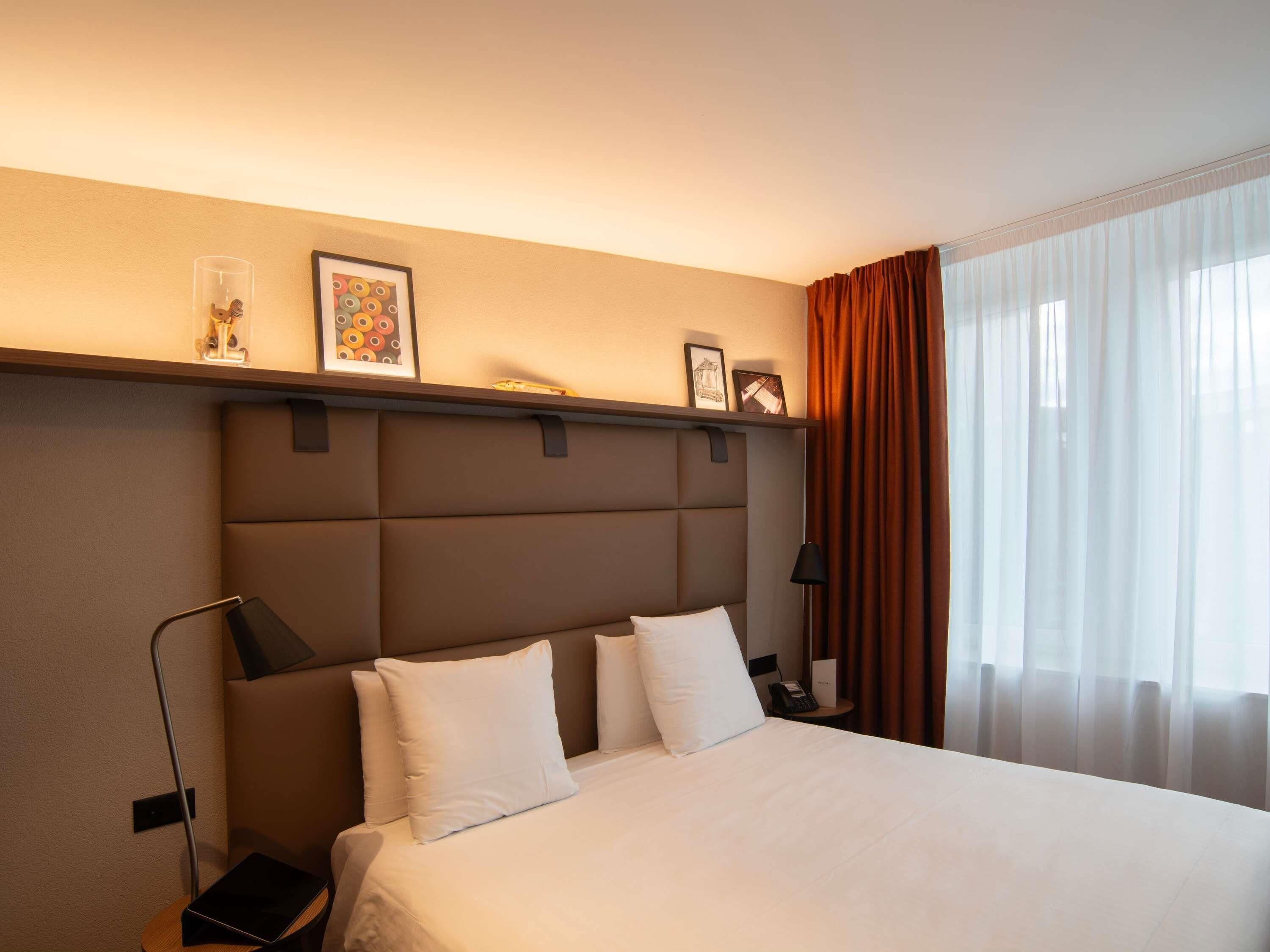 Quarto de hotel com uma cama grande, cabeceira, mesa de cabeceira com abajur, telefone e prateleira com quadros emoldurados.