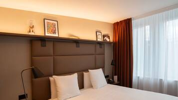 Quarto de hotel com uma cama grande, cabeceira, mesa de cabeceira com abajur, telefone e prateleira com quadros emoldurados.