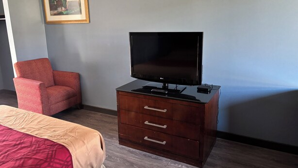Quarto de hotel com cômoda de madeira, televisão de tela plana, poltrona vermelha e cama com colcha vermelha e bege.