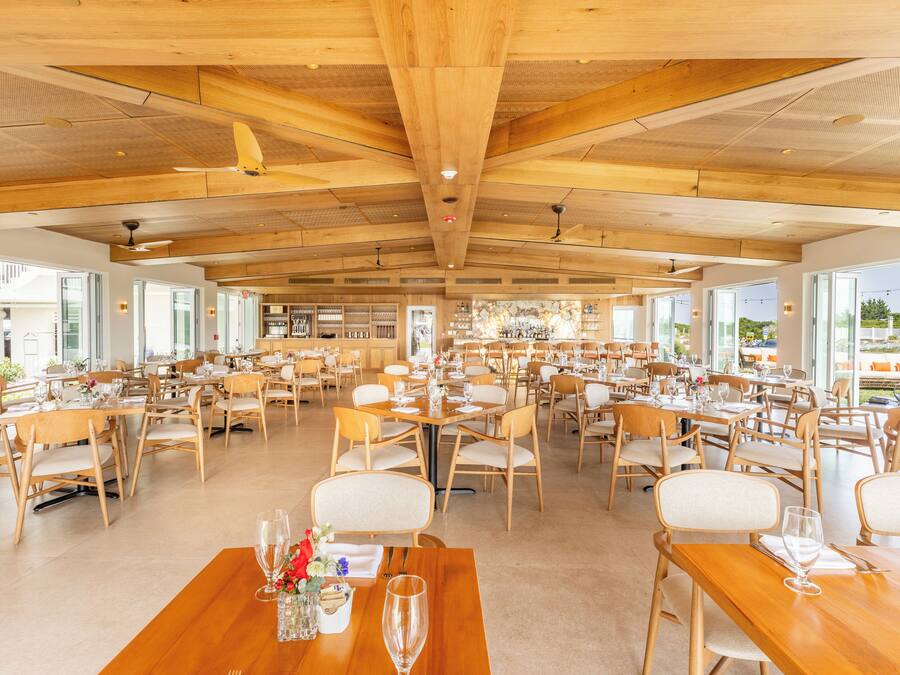Um restaurante amplo com tetos de madeira alta e grandes janelas que oferecem vista para o exterior. As mesas estão dispostas com cadeiras brancas e copos de vinho.