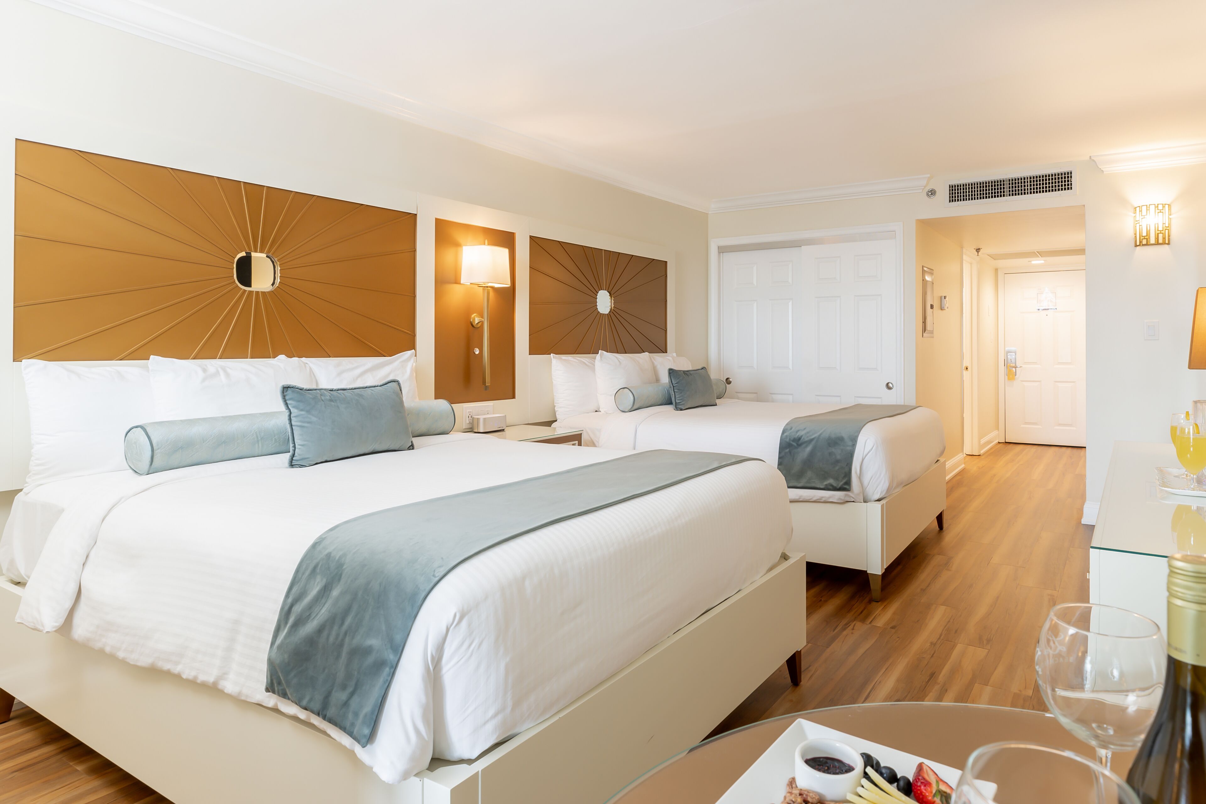 Quarto de hotel com duas camas, uma mesa de vidro com garrafa e copos, e uma porta que dá para outro quarto.
