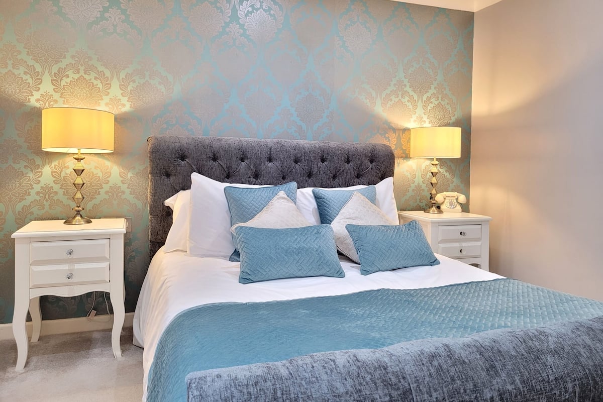 Quarto com cabeceira estofada, cama com roupa de cama azul, duas mesas de cabeceira com luminárias e um papel de parede estampado.