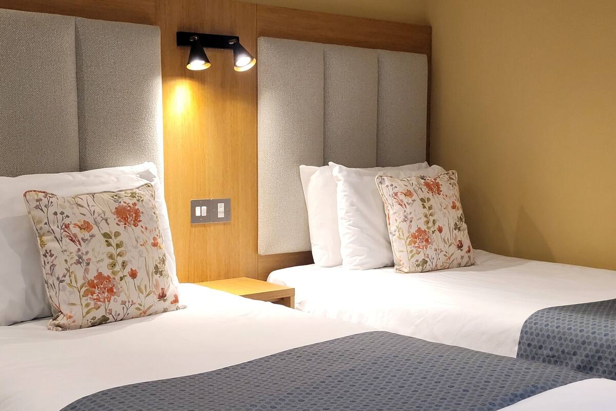 Quarto de hotel com duas camas, cada uma com roupa de cama branca e almofadas estampadas com flores. Possui cabeceira de madeira com iluminação embutida e uma pequena mesa de cabeceira.