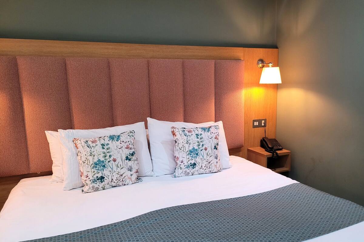 Quarto de hotel com uma cama grande, duas almofadas com padrão floral, cabeceira de madeira, luminária de cabeceira e telefone sobre uma mesinha de madeira.