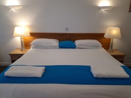 Cama bem arrumada com roupa de cama branca e azul, com duas luminárias laterais sobre mesas de cabeceira de madeira.