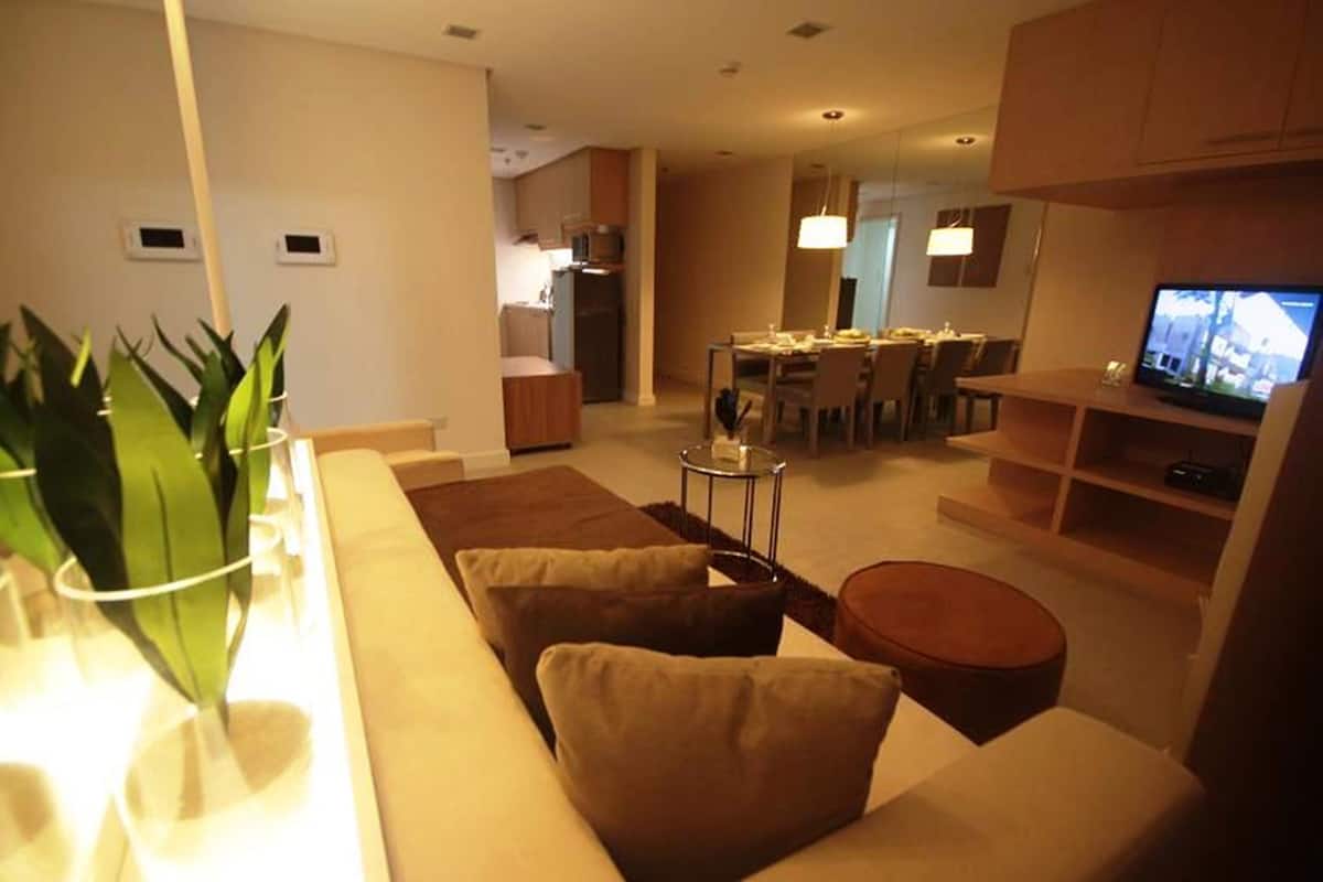 Uma sala de estar moderna com sofá, mesa de centro e televisão. Ao fundo, há uma área de jantar com mesa e cadeiras.