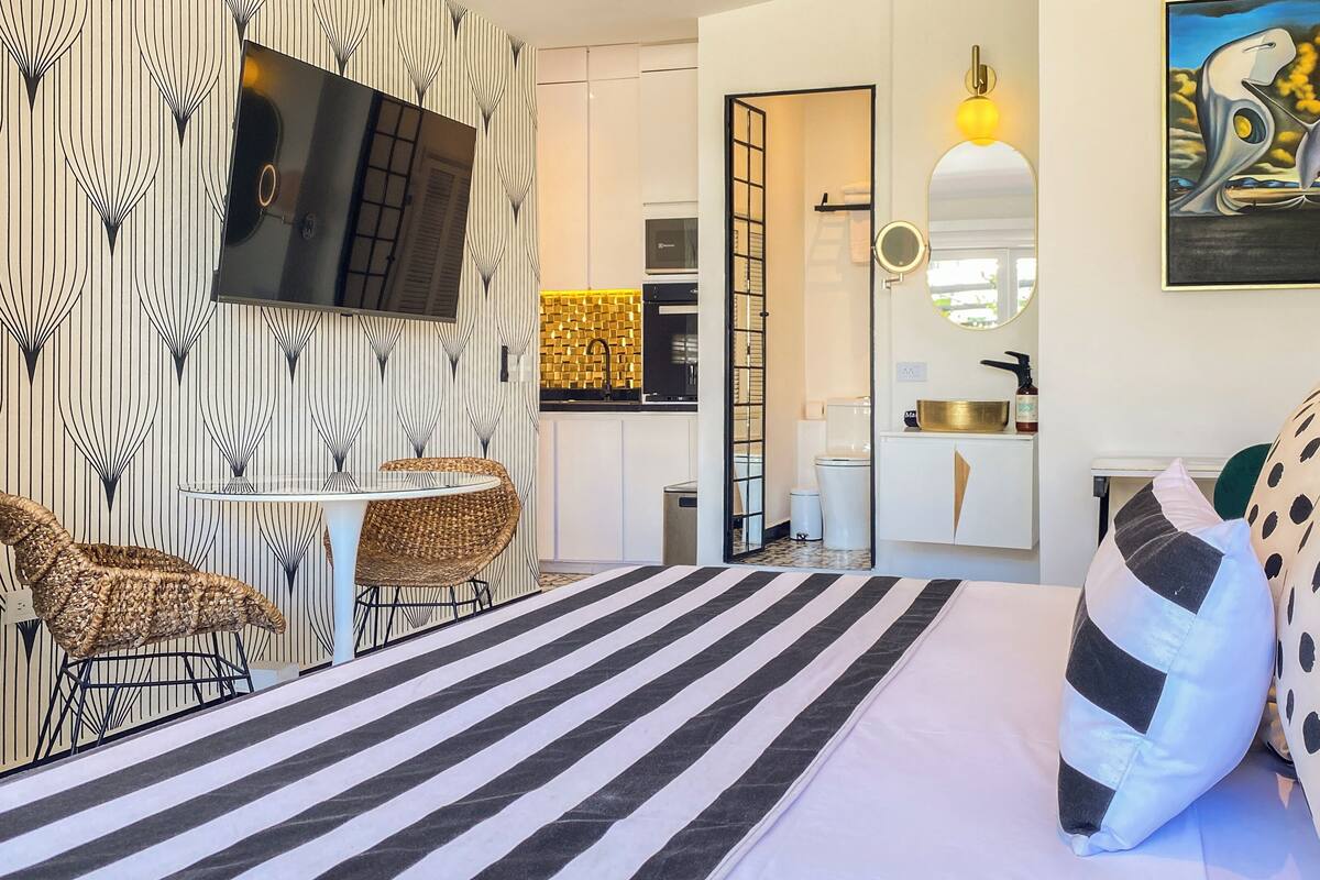 Quarto de hotel moderno com piso listrado, cama com travesseiro de bolinhas, televisão, área de refeições com cadeira de vime e cozinha visível por uma porta aberta.