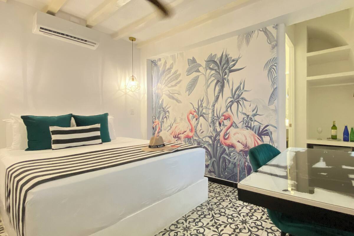 Quarto de hotel com cama, mesa, cadeira, parede com mural tropical e janela.