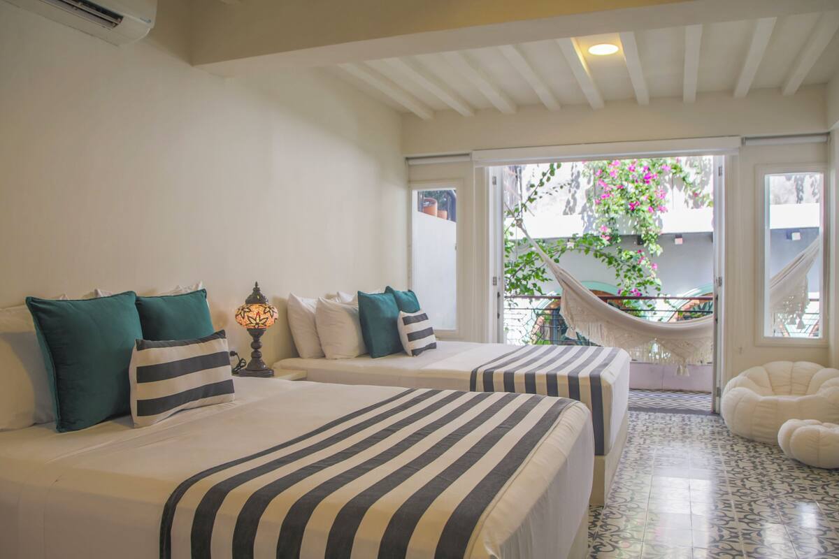 Quarto de hotel com duas camas, uma rede, varanda com plantas e uma cadeira branca.