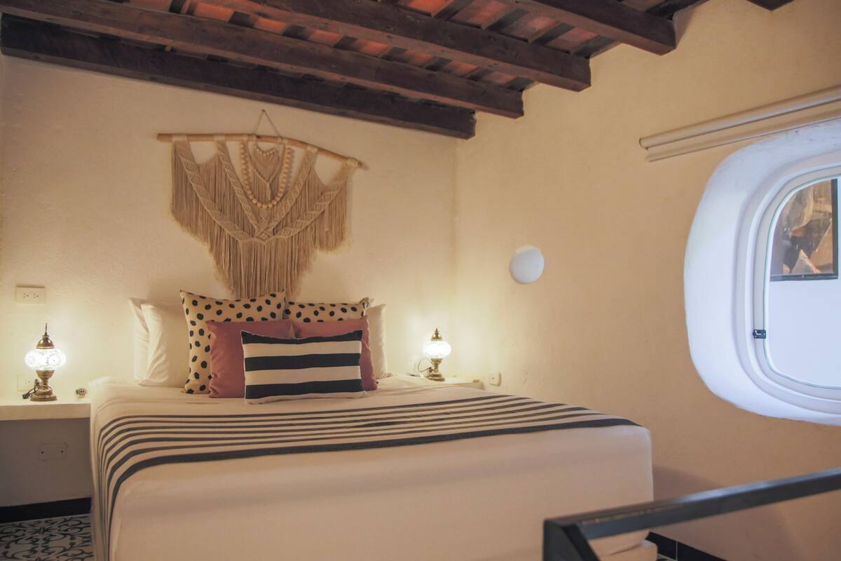 Quarto com cama, duas luminárias de cabeceira, um enfeite de macramê na parede e janela com vista.