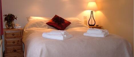 Cama bem arrumada com lençóis brancos e um travesseiro decorativo vermelho, uma luminária de cabeceira e uma pequena cômoda de madeira com gavetas.
