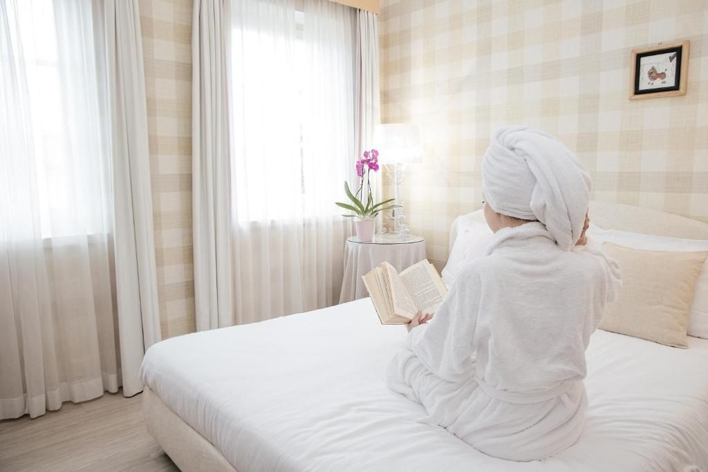 Uma pessoa de roupão branco lendo um livro em quarto com cama, uma mesinha com planta em vaso e janela com cortinas leves.
