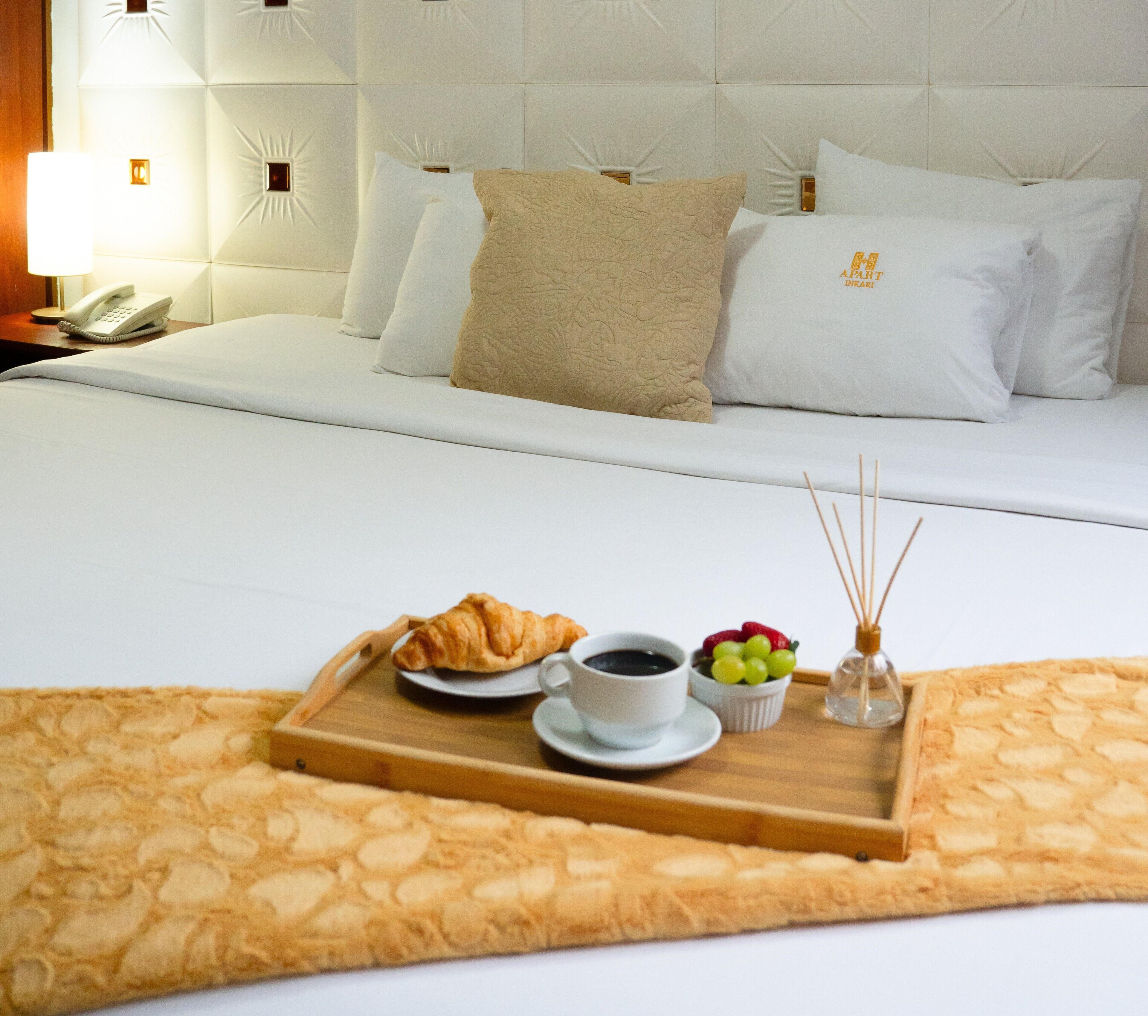 Quarto de hotel com uma cama bem arrumada, mesa de cabeceira com abajur, telefone e bandeja com croissant, café, frutas e um frasco de perfume.
