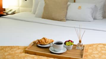 Quarto de hotel com uma cama bem arrumada, mesa de cabeceira com abajur, telefone e bandeja com croissant, café, frutas e um frasco de perfume.