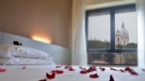 Quarto de hotel com cama, travesseiros e pétalas de rosa sobre a cama. Uma janela mostra um edifício com cúpula ao fundo.