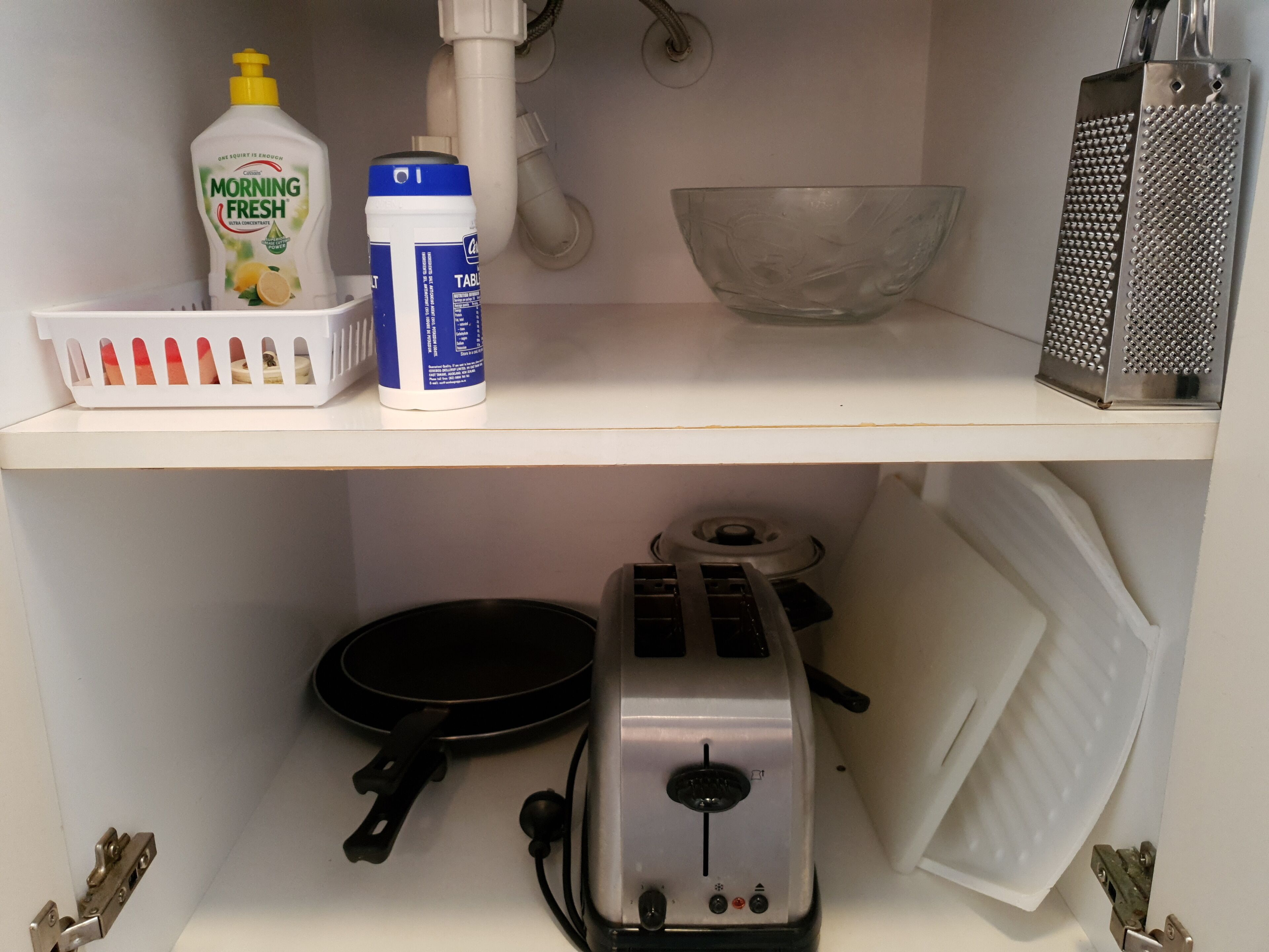 Uma prateleira na cozinha com um frasco de detergente, um recipiente de bicarbonato de sódio, um ralador e uma frigideira.