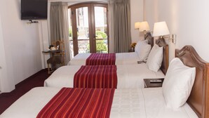 Quarto de hotel com duas camas, uma televisão na parede, uma mesinha com uma cadeira e vista para o exterior por uma porta de vidro.