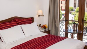Quarto de hotel com uma cama grande, uma mesinha de cabeceira com abajur, um vaso com flores e uma varanda com vista para a rua.