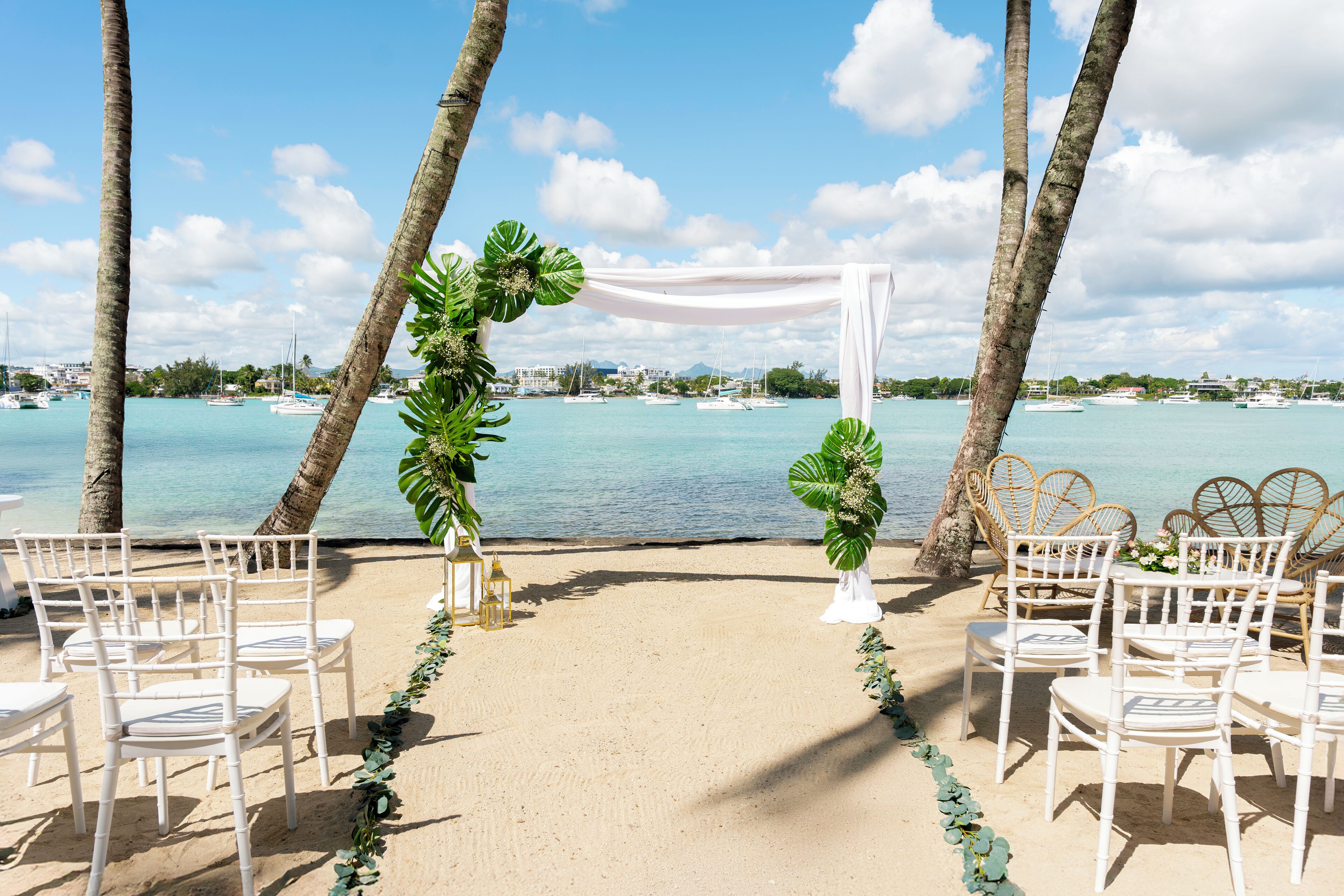 Preparação para um casamento na praia, com cadeiras brancas, um toldo, palmeiras e vista para o mar e barcos.