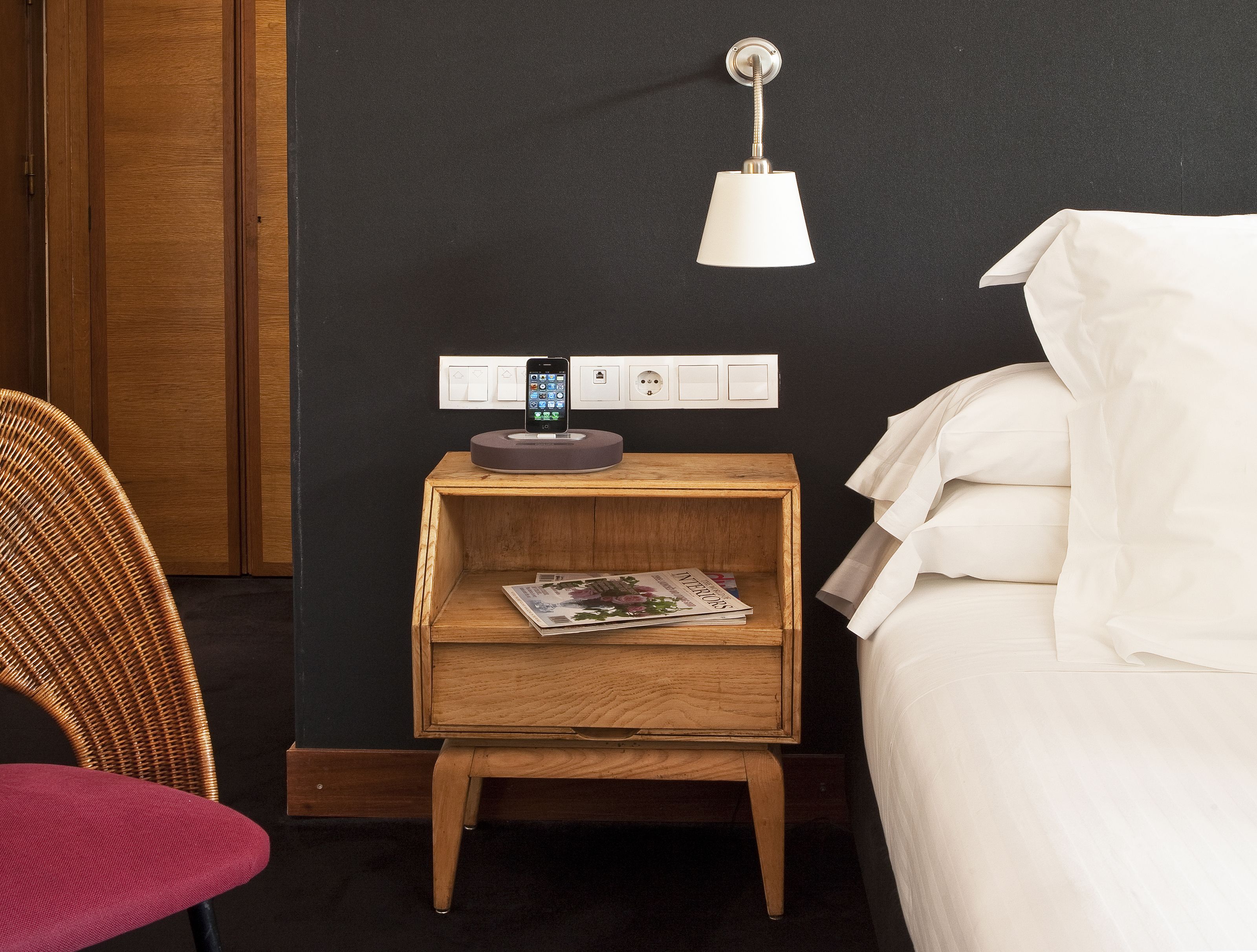 Quarto de hotel com mesa de cabeceira de madeira, luminária, base de carregamento para smartphone, revista, cadeira de vime e cama com roupa de cama branca.