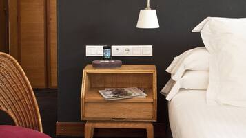 Quarto de hotel com mesa de cabeceira de madeira, luminária, base de carregamento para smartphone, revista, cadeira de vime e cama com roupa de cama branca.