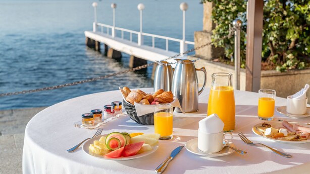 Mesa de pequeno-almoço com croissants, fruta e bebidas, com vista para o lago.