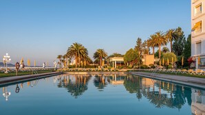 Piscina com reflexos límpidos, rodeada por palmeiras e espreguiçadeiras, com um edifício ao fundo.