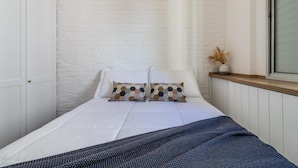 Cama bem arrumada com roupa de cama branca e almofada estampada, parede de tijolos brancos e janela com vista para o exterior.