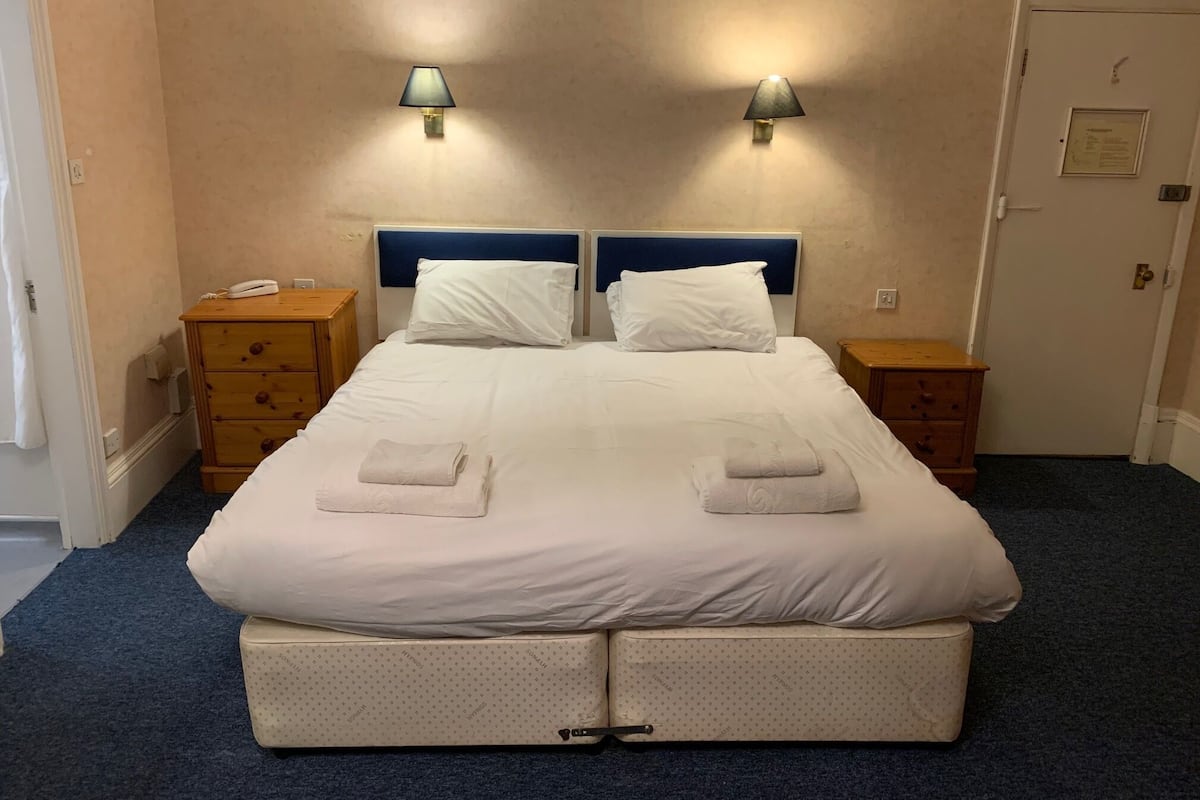 Uma cama de casal com duas mesas de cabeceira, um encosto e duas luminárias fixadas na parede.