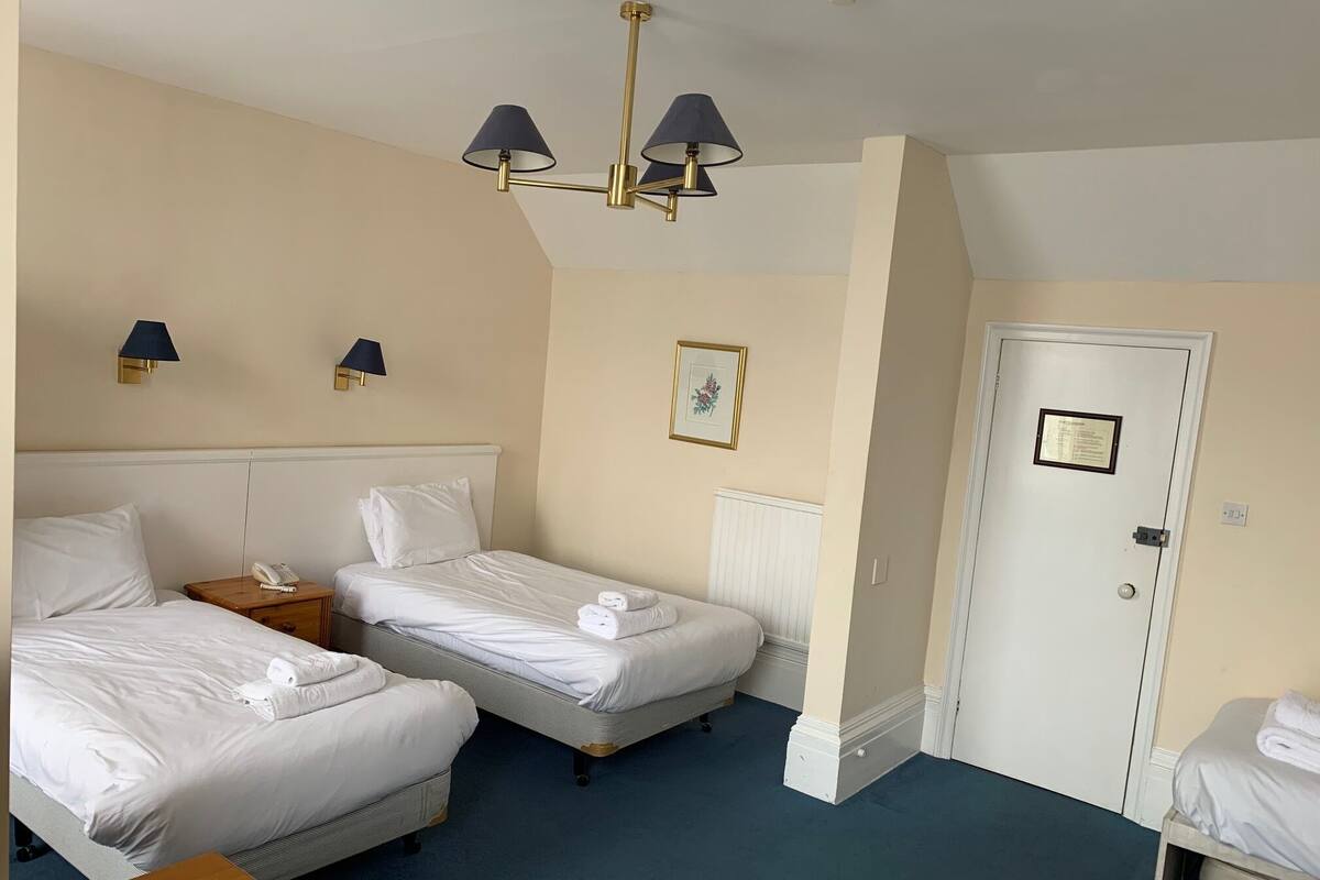 Quarto de hotel com duas camas de solteiro, uma mesa de cabeceira de madeira com telefone, uma pequena mesa, um lustre e uma porta com placa.