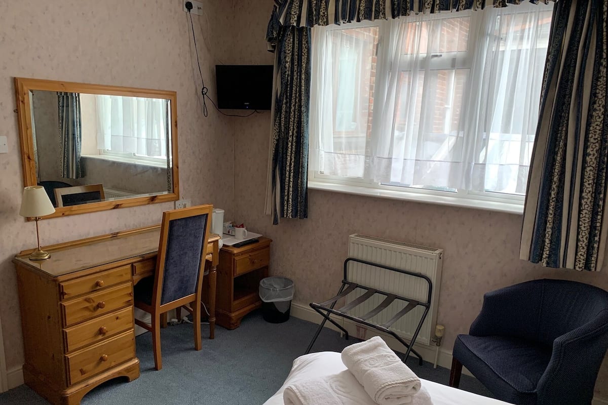 Quarto de hotel com penteadeira de madeira, escrivaninha com cadeira, cama com toalhas dobradas, radiador, televisão fixada na parede e janela com cortinas.