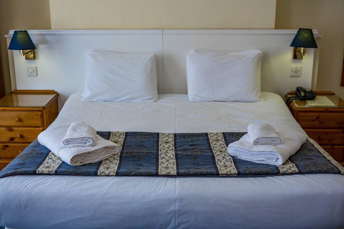 Cama bem arrumada com lençóis brancos e edredom estampado em azul e branco. Há duas mesas de cabeceira com abajures, um aparador de madeira e um telefone em uma das mesas.