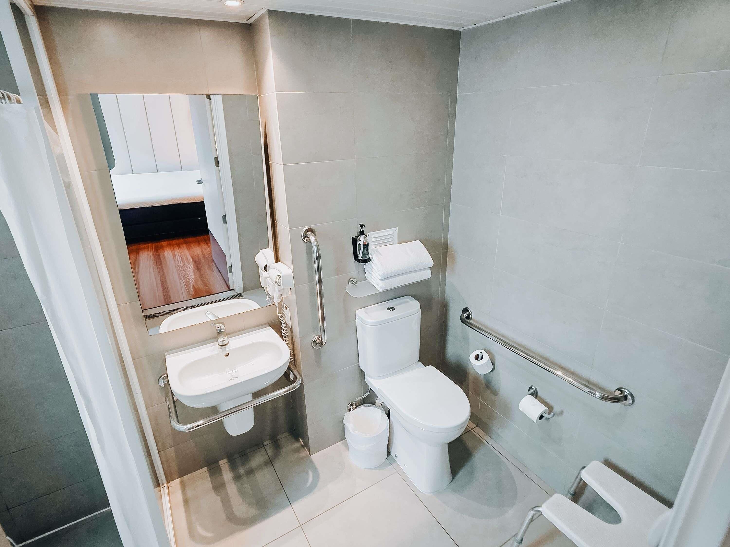 Casa de banho moderna com lavatório, sanita e bidé, equipada com barras de apoio e espelho.