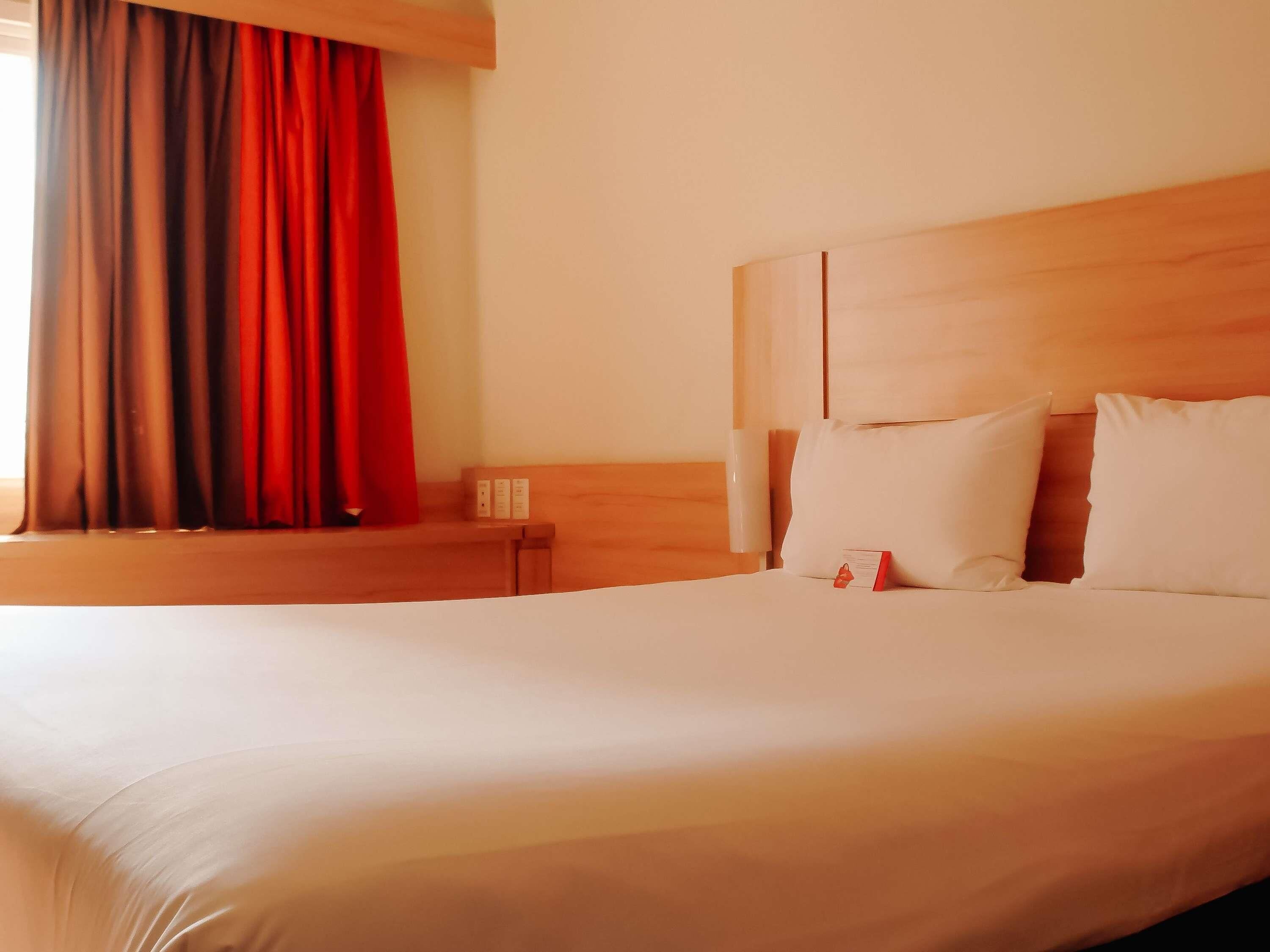Quarto de hotel com uma cama grande, cabeceira de madeira, almofadas brancas, mesa de cabeceira com abajur e uma janela com cortinas vermelhas.