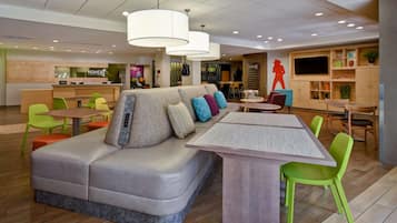 Um hall moderno de hotel com área de estar, apresentando um sofá cinza com diversas almofadas, uma mesa de recepção de madeira e uma grande TV de tela plana fixada em um móvel de madeira.