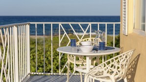 Varanda com mesa e cadeiras brancas, uma tigela, um jarro e copos empilhados. A vista inclui o oceano e vegetação.
