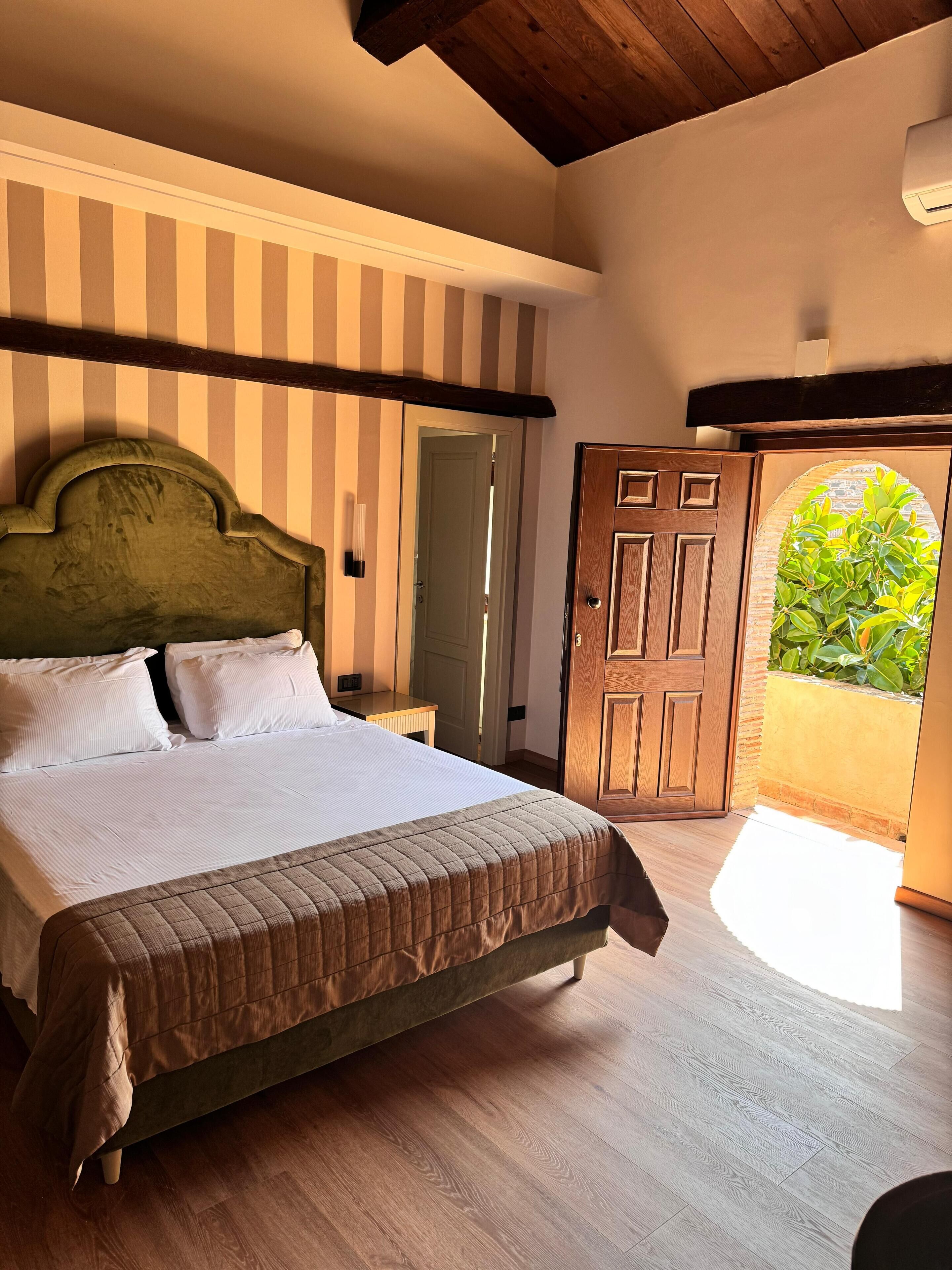Quarto com cama grande, cabeceira de madeira e vista para um jardim através de uma porta aberta.