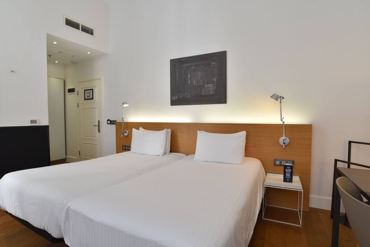 Quarto de hotel com duas camas, cabeceira de madeira, obra de arte na parede, mesa de cabeceira com abajur e pequena escrivaninha com cadeira.