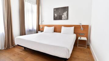 Quarto de hotel com uma cama grande, cabeceira de madeira, mesa de cabeceira pequena, luminária na parede e um quadro.