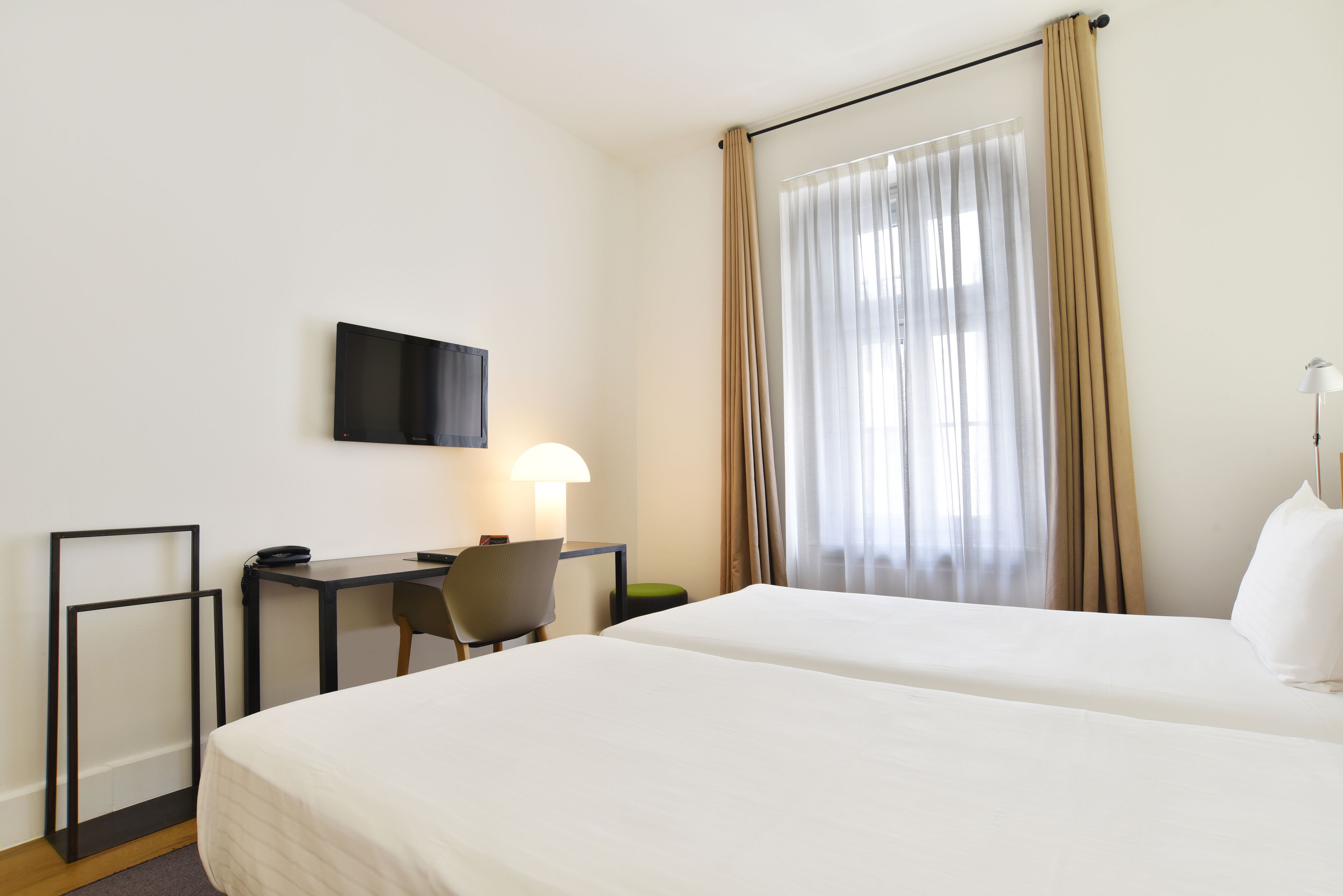 Quarto de hotel com cama, escrivaninha, cadeira, televisão e janela com cortinas.